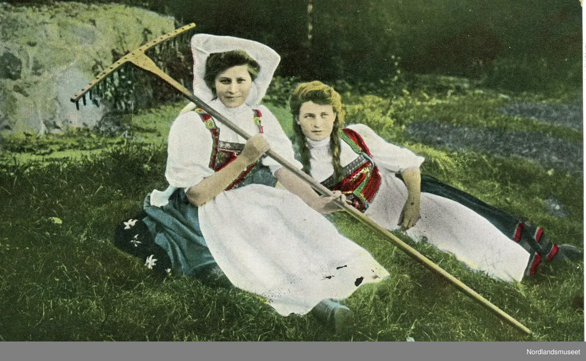 Postkort med bilde av to kvinner i bunad som ligger på en gresseng. Den ...