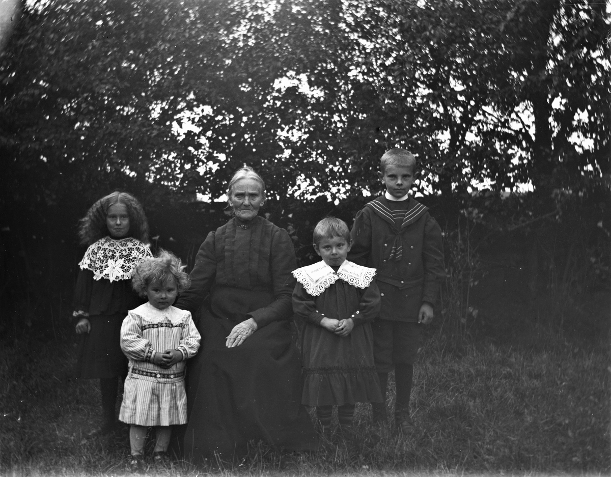 En äldre kvinna sitter utomhus med fyra barn. Pojkarna har olika koltar ...