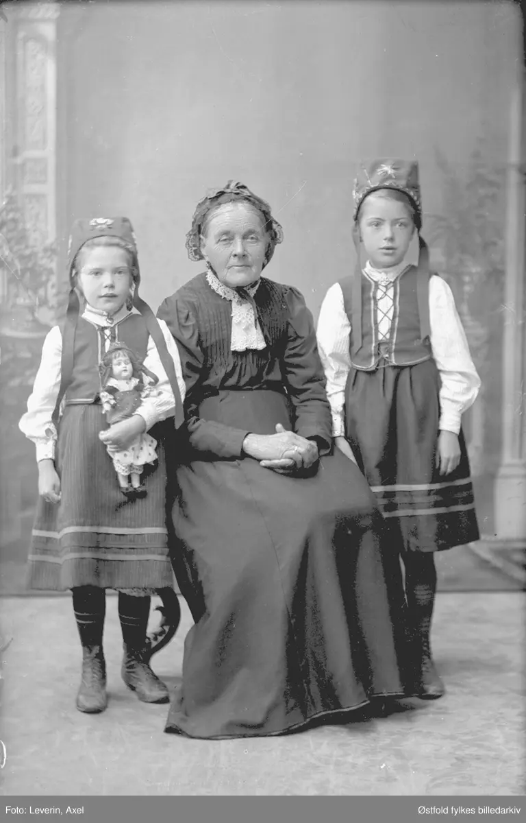 Ukjent eldre kvinne (bestemor?) med to barn, jenter/søstre i bunad ...