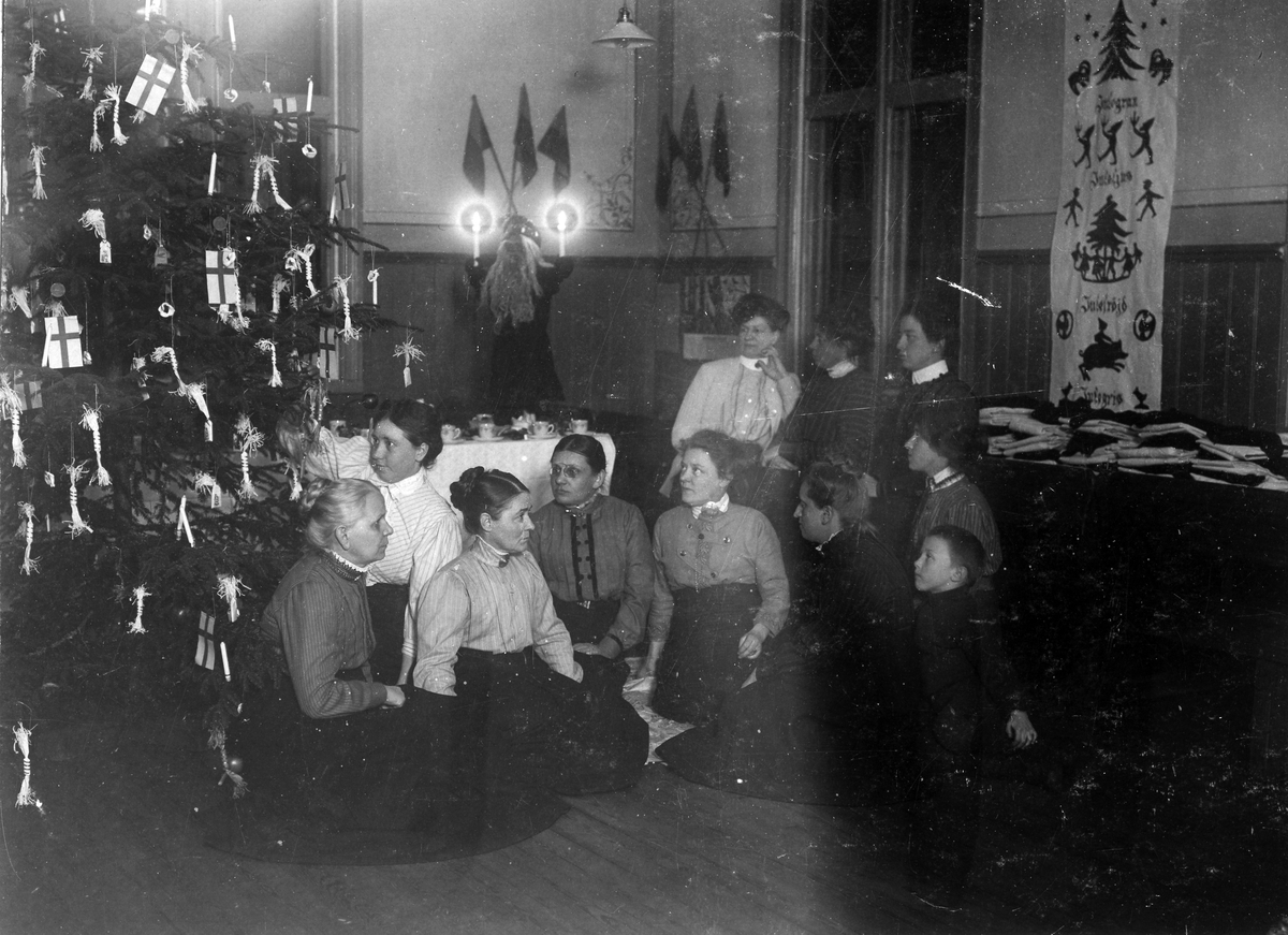 10 kvinnor och pojke sitter i ett julpyntat rum. Till vänster en pyntad ...