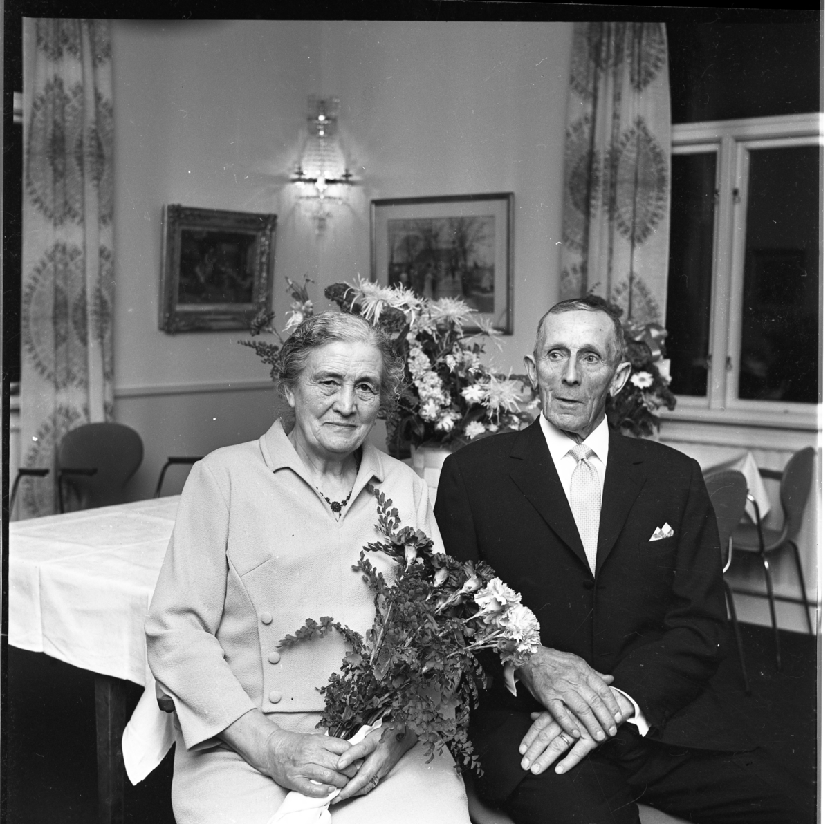 Ett äldre par sitter i sitt hem, kvinnan har en blomsterbukett i handen ...