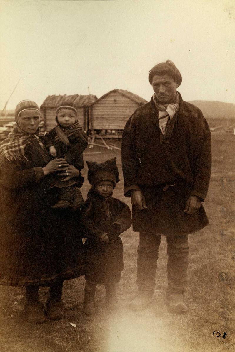 Norge. Mann, kvinne og to barn i samisk drakt, står foran to tømmerhus ...