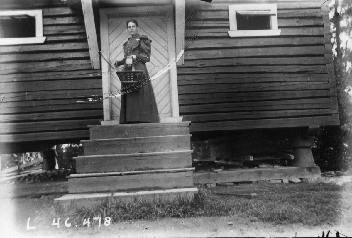 Drakt 1908-10. Kvinne stående på en trapp. I bakgrunnen et stabbur ...
